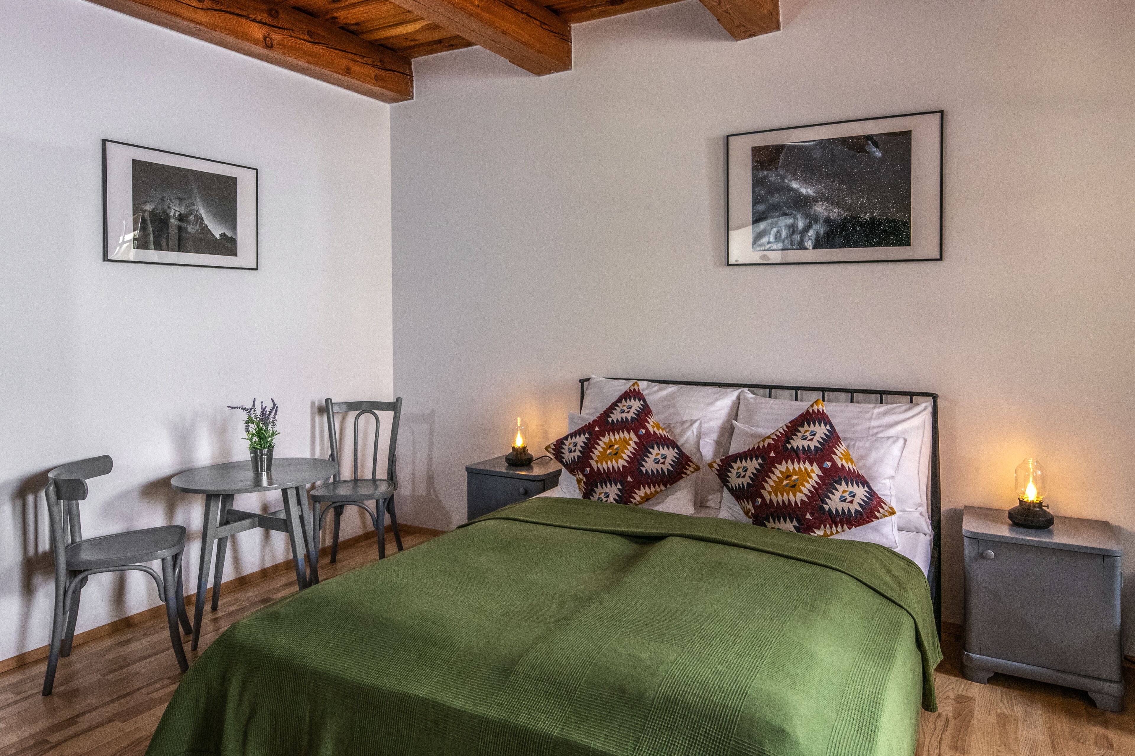 Quarto com uma cama, duas mesas de cabeceira com luminárias, uma mesinha com cadeiras e duas molduras com quadros na parede.