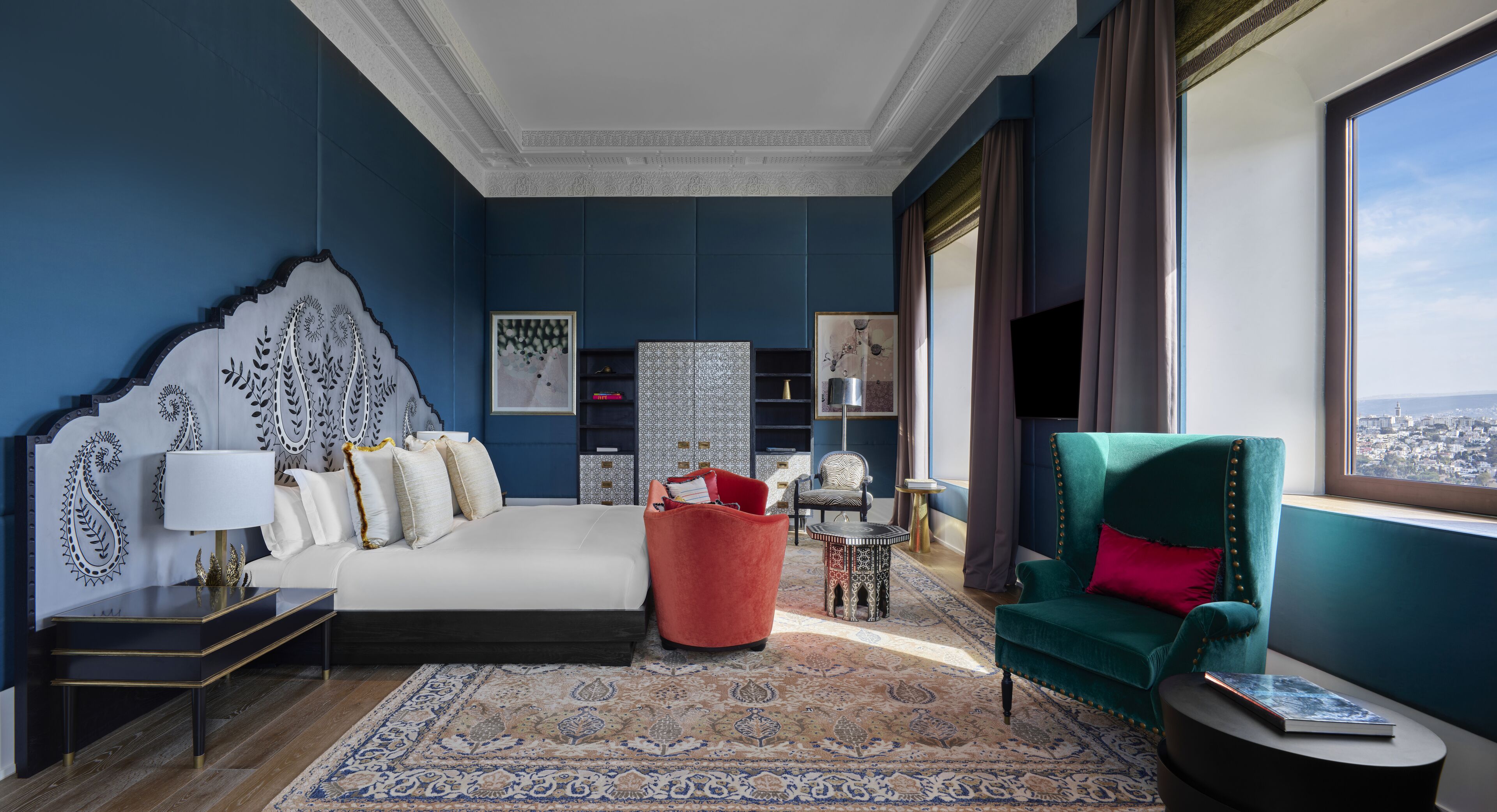 Um quarto moderno com uma cama grande, uma poltrona vermelha, uma poltrona verde-água, uma pequena mesa redonda e vista da cidade através de uma grande janela.