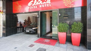Entrada do Hotel Gloria Plaza, com toldo vermelho, um carro preto estacionado e duas grandes plantas em vasos à frente.