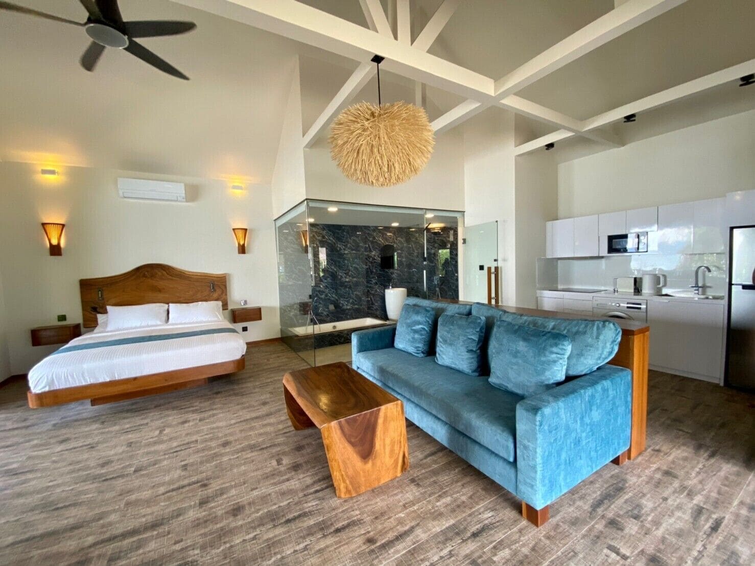 Quarto de hotel moderno com cama, sofá azul, mesa de centro de madeira, banheiro com divisória de vidro e cozinha compacta.