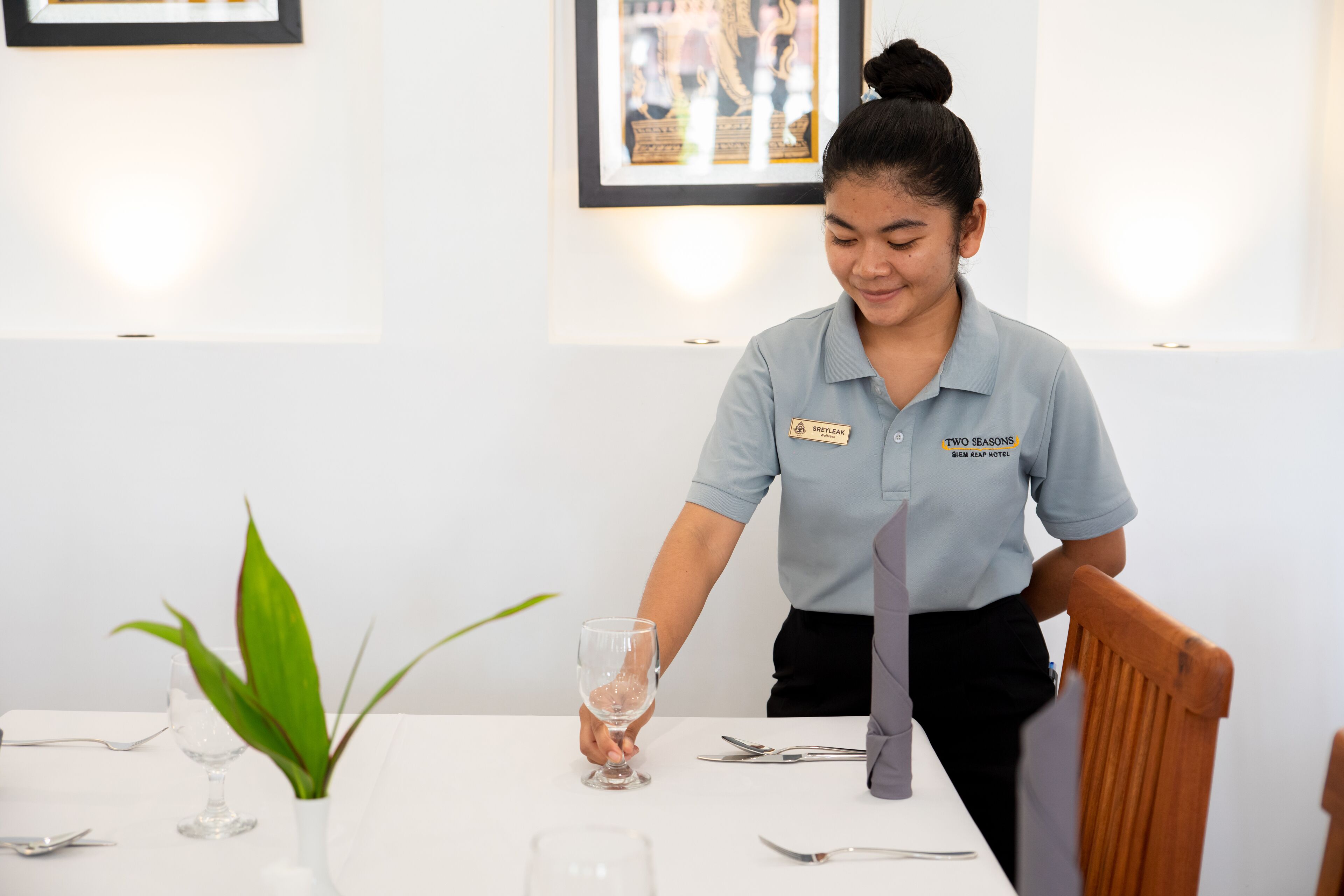 Um funcionário de hotel sentado à mesa, com uma placa de identificação, um copo e uma planta.