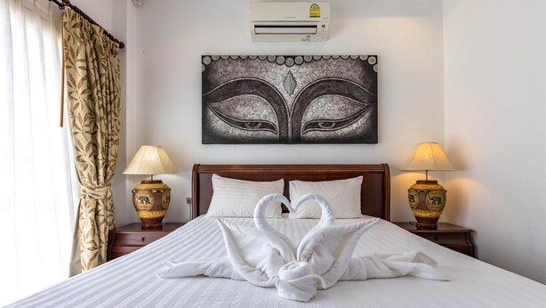 Cama bem arrumada com roupa de cama branca e arranjo de toalhas em formato de coração. Há duas luminárias de cabeceira com vasos decorativos, um ar condicionado fixado na parede e uma grande pintura abstrata sobre a cama.