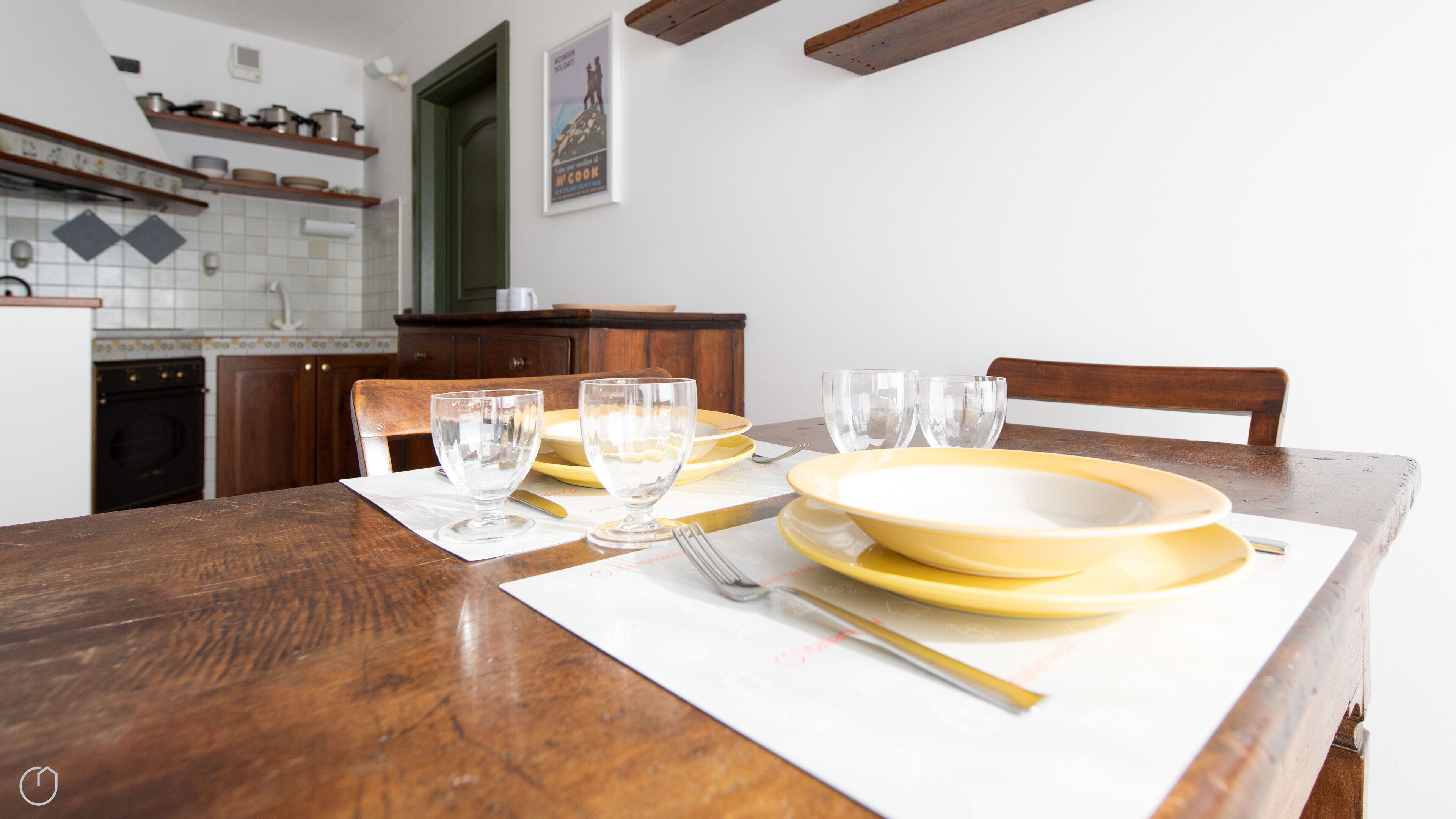 Mesa de jantar posta com pratos amarelos, copos transparentes e um garfo sobre um jogo americano branco.