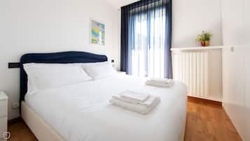 Cama bem arrumada com roupa de cama branca e toalhas dobradas, cabeceira azul escura, janela com cortinas azuis, uma pequena planta em vaso sobre um prateleira branca, e um radiador na parede.