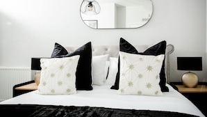 Cama bem arrumada com edredom preto e branco, dois travesseiros pretos e dois travesseiros brancos com estampas de estrelas. Um espelho redondo pendurado acima da cama, e um abajur sobre uma mesa de cabeceira.