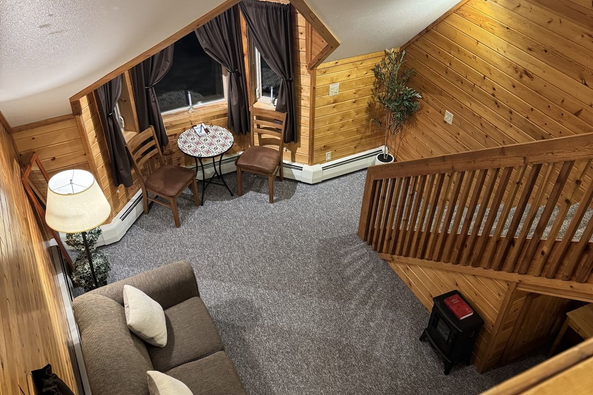 Um quarto acolhedor estilo loft, com teto de madeira, sofá com almofadas, mesa pequena com cadeiras e janela com cortinas.