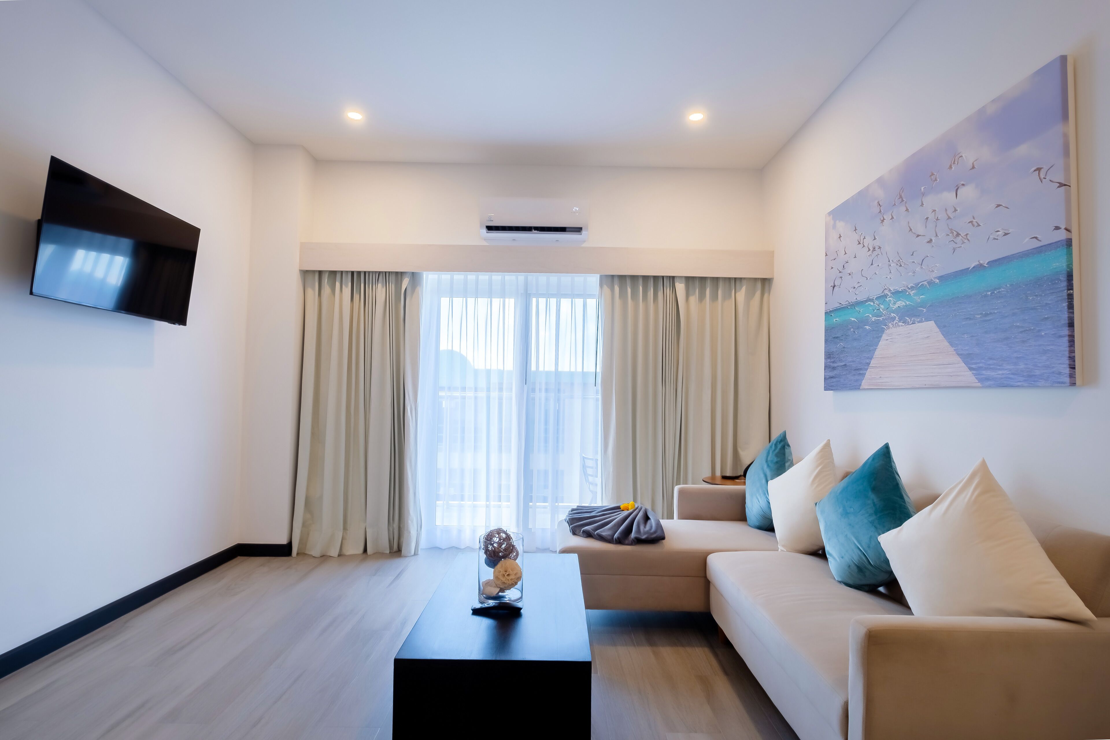 Uma sala de estar moderna com uma televisão de tela plana, uma janela grande com cortinas, um sofá bege com almofadas verde-água e uma pintura de um cais e gaivotas.