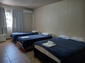 Quarto com duas camas, cada uma com um travesseiro e uma toalha enrolada. Há uma janela com cortinas, um ar condicionado e uma mesa pequena com abajur.