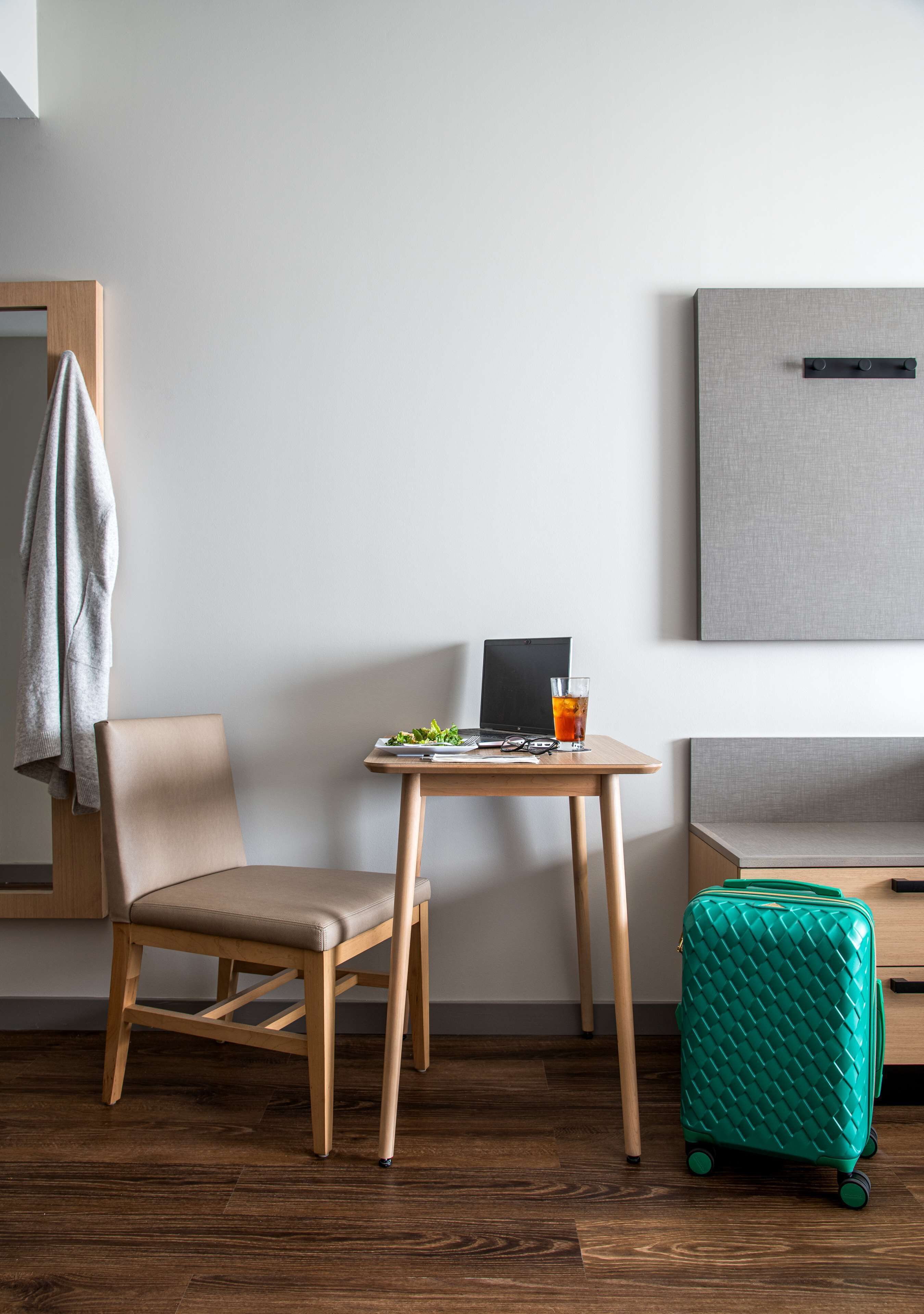 Quarto moderno com uma cadeira de madeira, uma mesa pequena com um laptop e uma bebida, e uma mala verde-água.