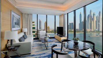 Quarto de hotel moderno com grande janela e vista para o horizonte urbano, um sofá, uma mesa de jantar e uma televisão.