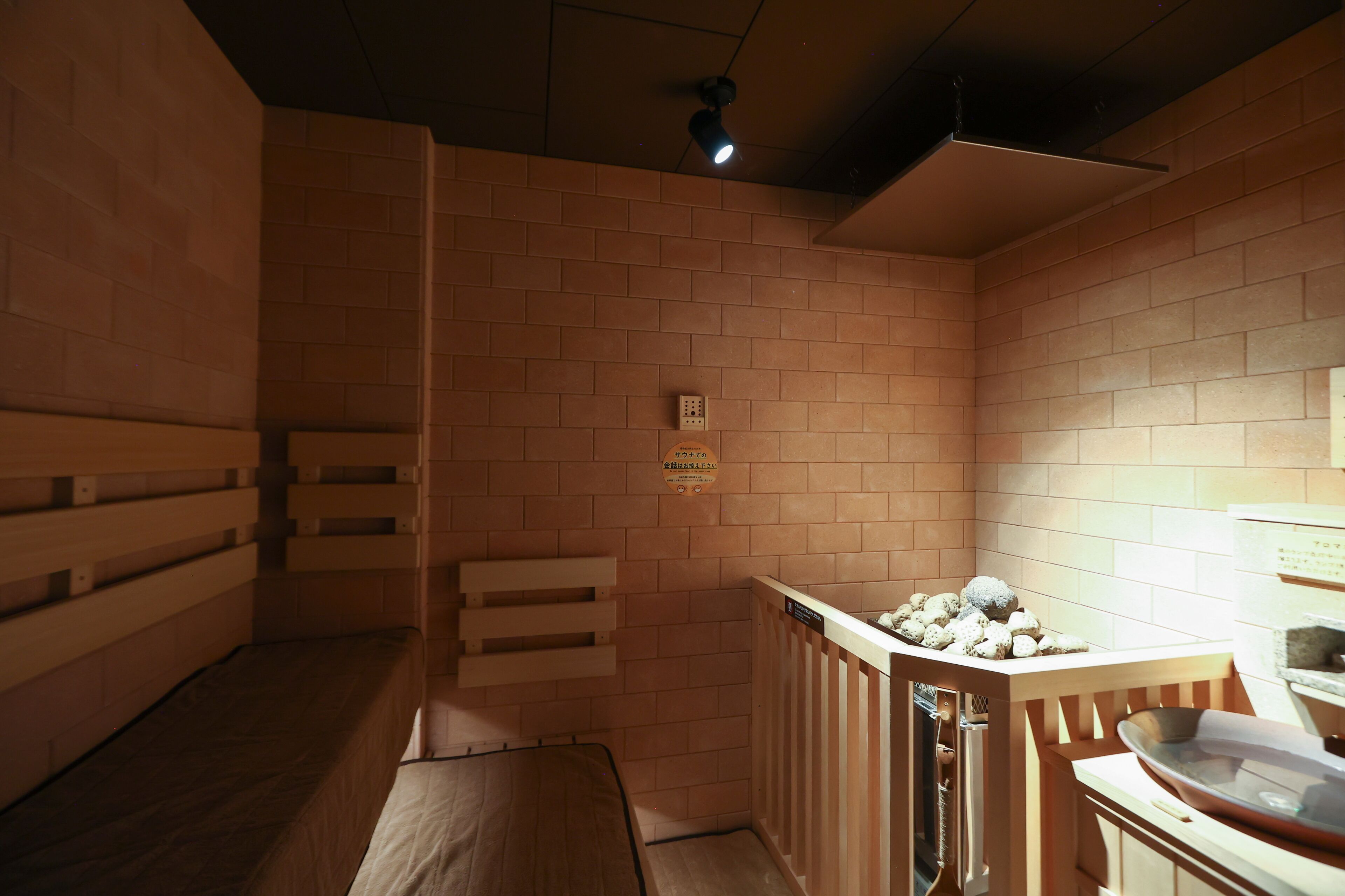 Uma sala de sauna com bancos de madeira, um banco na entrada, uma pequena mesa com uma tigela de pedras e uma parede revestida de azulejos.