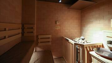 Uma sala de sauna com bancos de madeira, um banco na entrada, uma pequena mesa com uma tigela de pedras e uma parede revestida de azulejos.