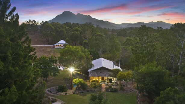 Uma casa de campo com fachada de pedra e telhado de colmo, rodeada por uma floresta densa e uma montanha ao fundo.
