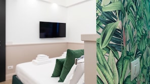 Quarto de hotel com cama, almofadas verdes, televisão de tela plana na parede e papel de parede tropical.