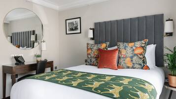 Quarto de hotel com uma cama grande, uma escrivaninha com um laptop, um espelho, um quadro na parede e uma planta em vaso.