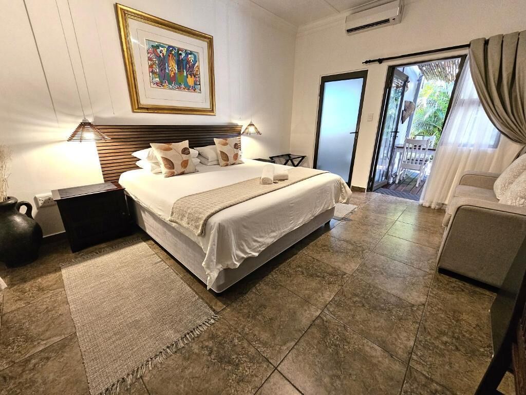 Quarto de hotel com uma cama, mesa de cabeceira, luminária, uma janela grande e porta que dá para uma varanda.