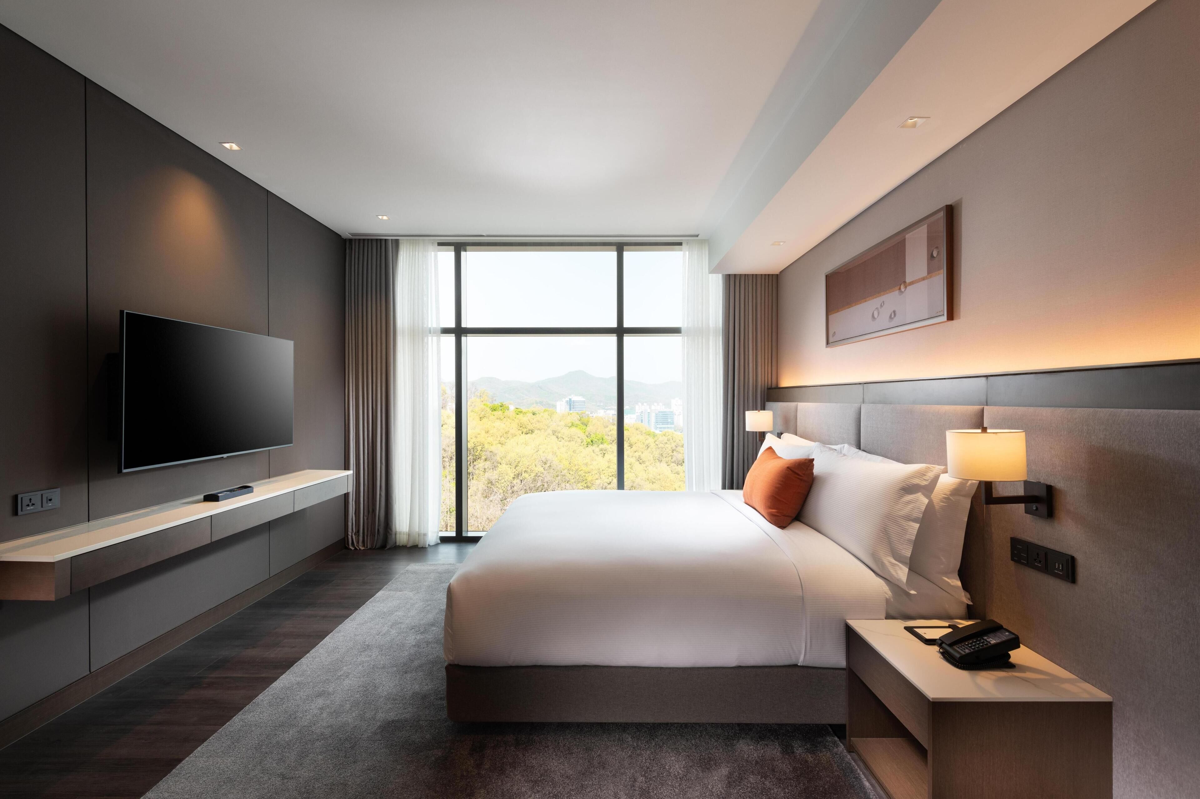 Quarto de hotel moderno com uma cama grande, televisão de tela plana, mesa de cabeceira com abajur e vista para o exterior.