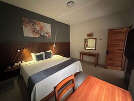 Quarto de hotel com cama, mesa de cabeceira, cadeira de madeira, mesa e espelho na parede.