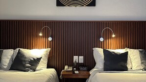 Quarto de hotel com duas camas, parede revestida de painel de madeira, obra de arte pendurada na parede, luminárias de cabeceira, mesa de cabeceira com telefone e relógio, e piso ladrilhado.