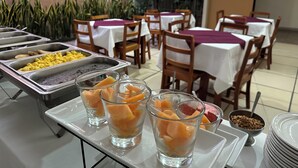 Uma área de buffet com diversas opções de comida, incluindo frutas e cereais, e uma televisão fixada na parede.