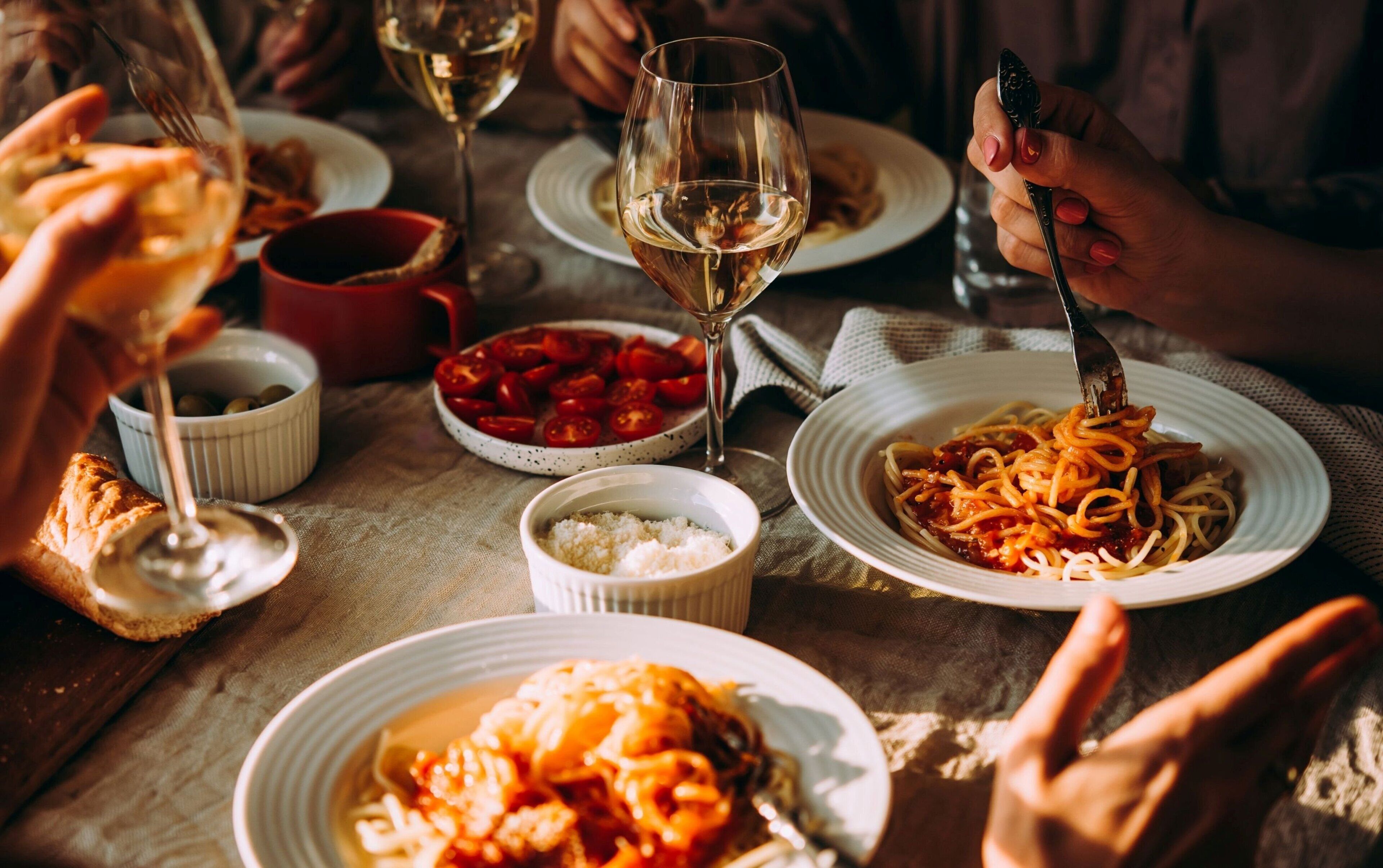 Uma mesa de jantar com pratos de comida, copos de vinho e uma pessoa usando um garfo.