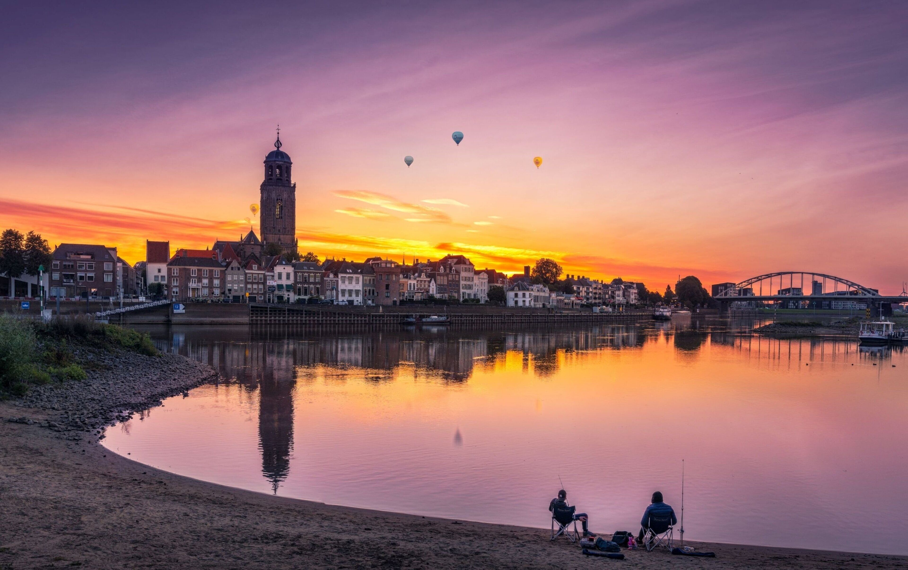 Uma paisagem urbana à beira-mar, com uma torre imponente, águas calmas refletindo os edifícios e uma ponte ao longe.