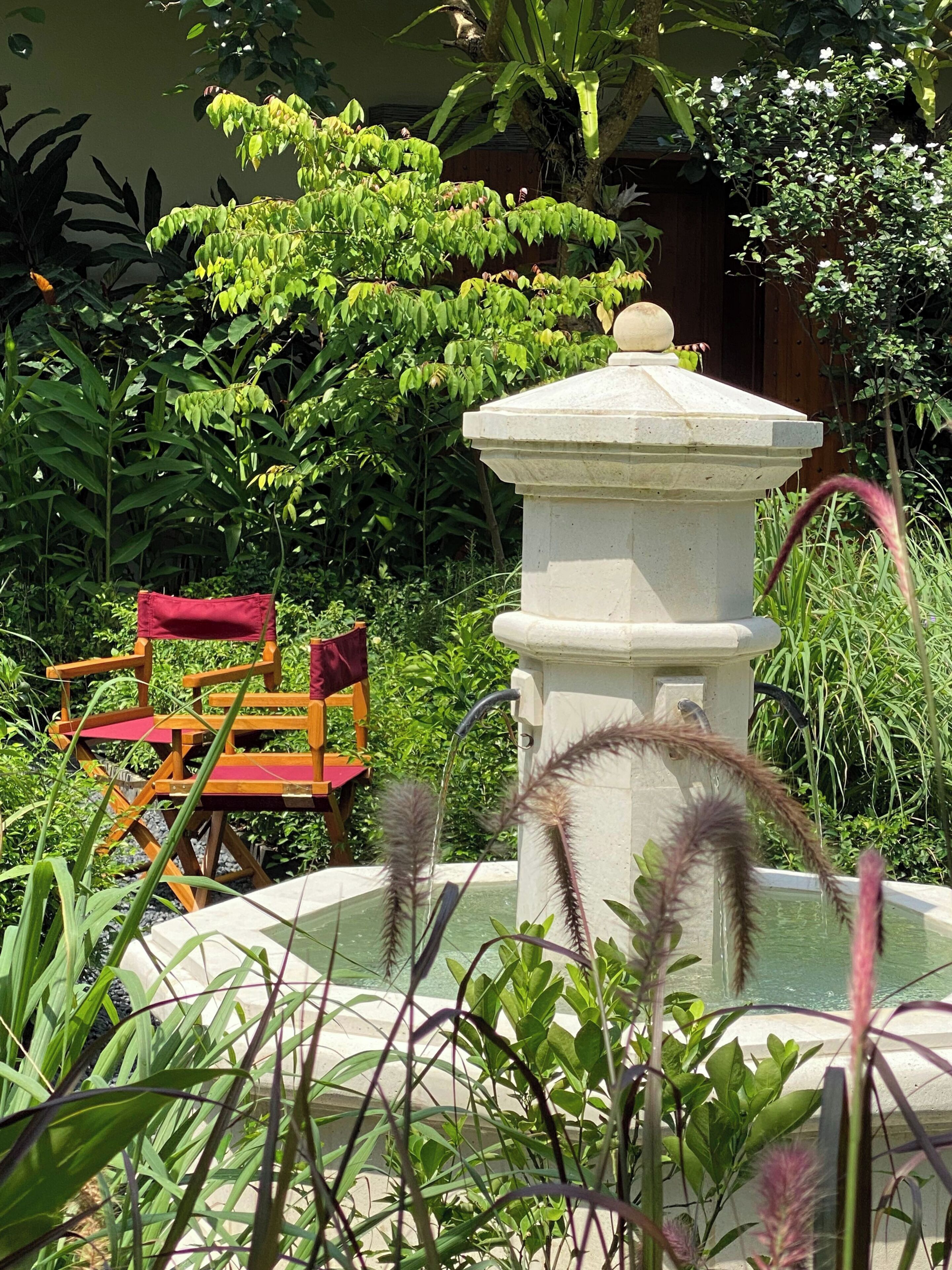 Um jardim com uma fonte, uma cadeira de madeira e vegetação exuberante.