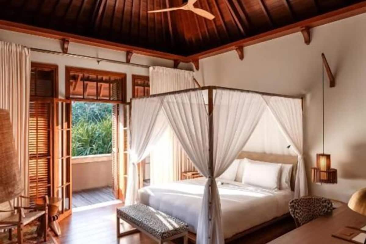 Quarto com cama, um banco, uma mesa, luminária pendente e vista para o verde lá fora.