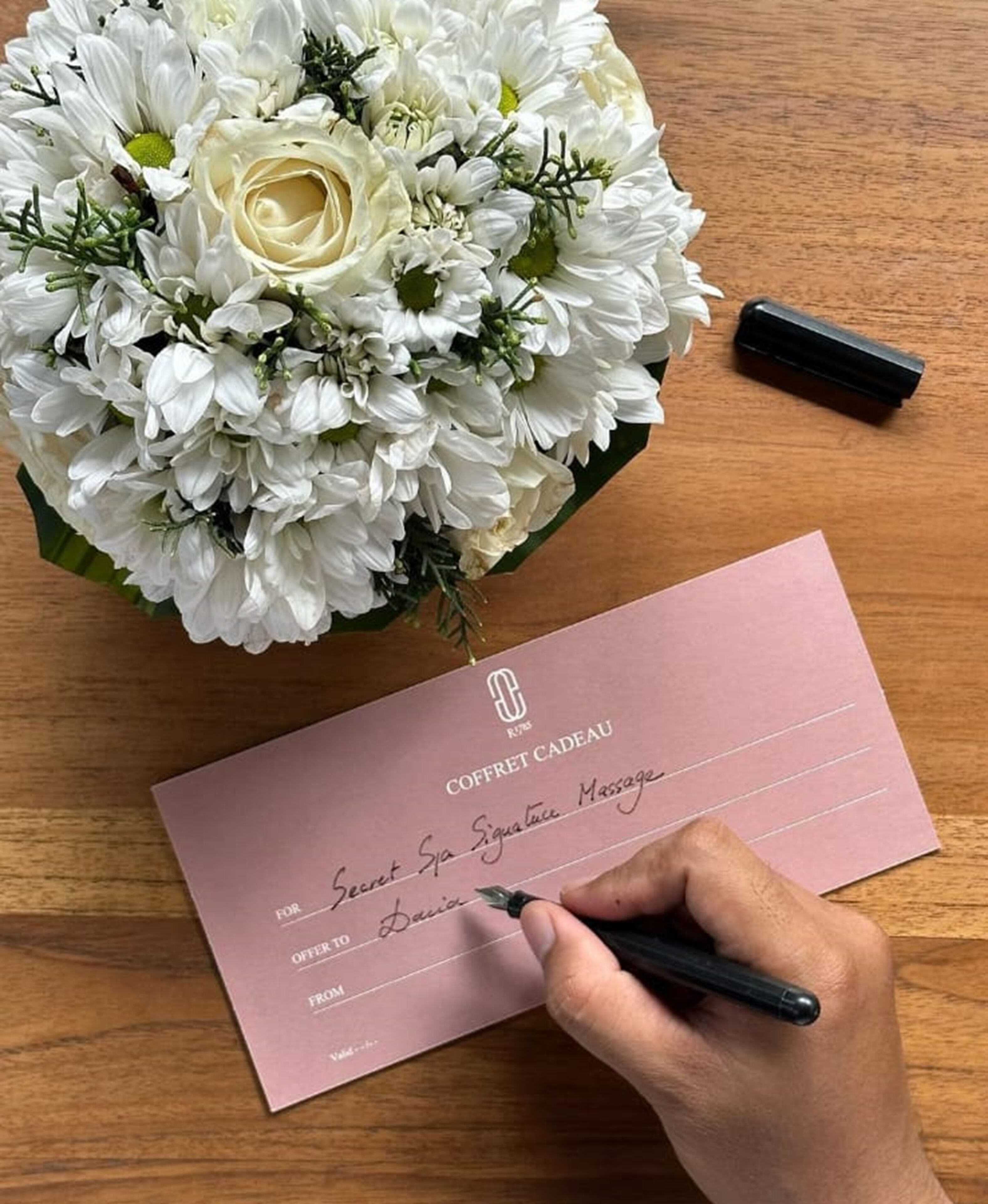 Uma mão escrevendo em um cartão rosa com uma caneta preta, colocado sobre uma mesa de madeira ao lado de um buquê de flores brancas.