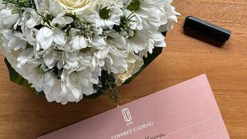 Uma mão escrevendo em um cartão rosa com uma caneta preta, colocado sobre uma mesa de madeira ao lado de um buquê de flores brancas.