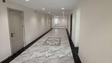Um corredor longo e estreito com paredes brancas e chão de mármore, que leva a uma porta no final.