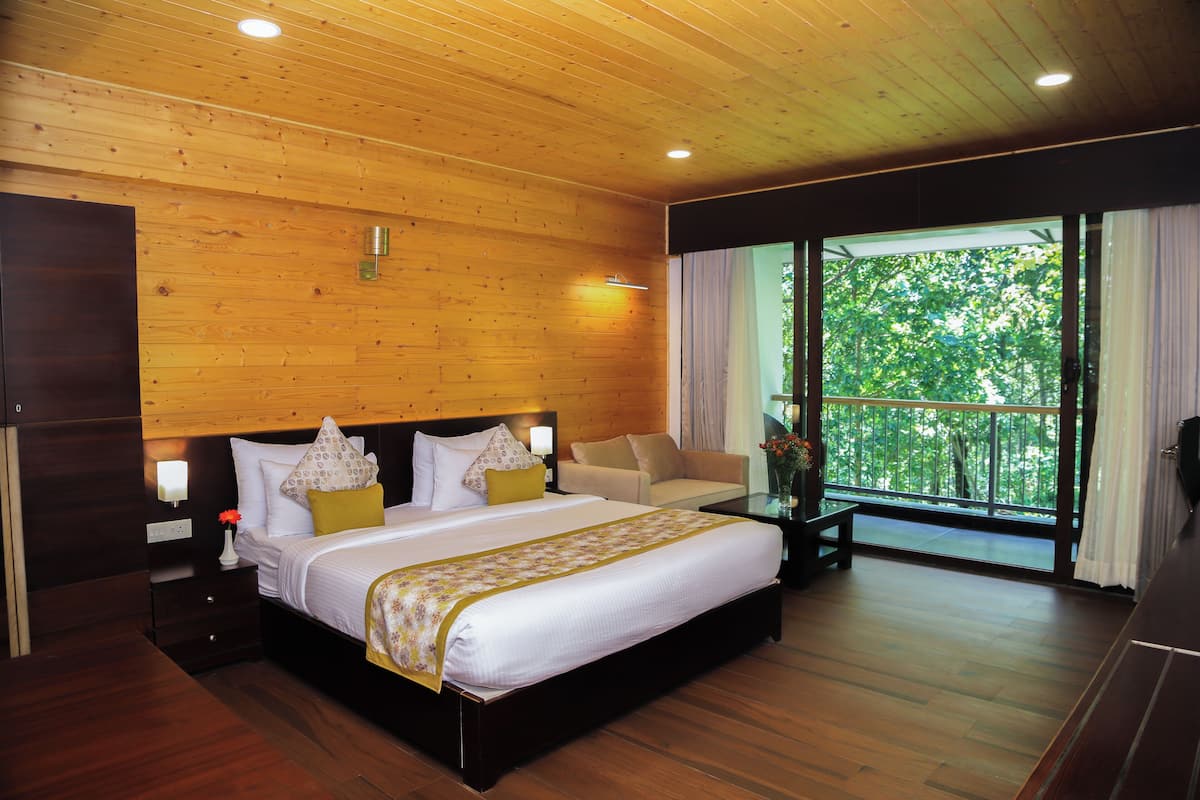 Quarto de hotel com teto de madeira, uma cama grande com colcha florida, um sofá, uma mesa de centro de vidro e vista para o verde através de uma janela ampla.