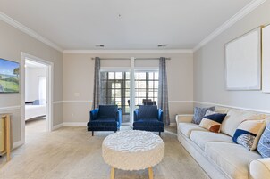 Uma sala de estar com carpete bege, um banquinho redondo, um sofá branco, duas poltronas azuis e uma janela grande com cortinas.