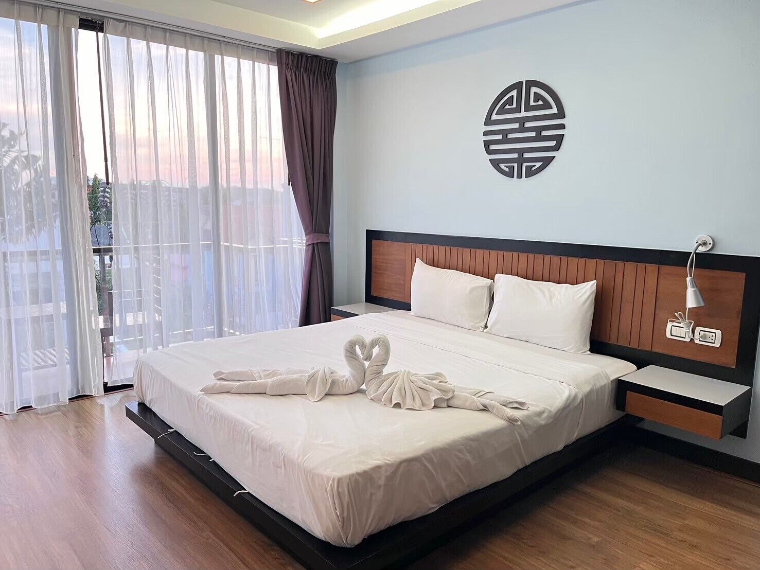 Quarto de hotel com uma cama grande, cabeceira de madeira, luz acoplada à parede e vista através de cortinas finas.