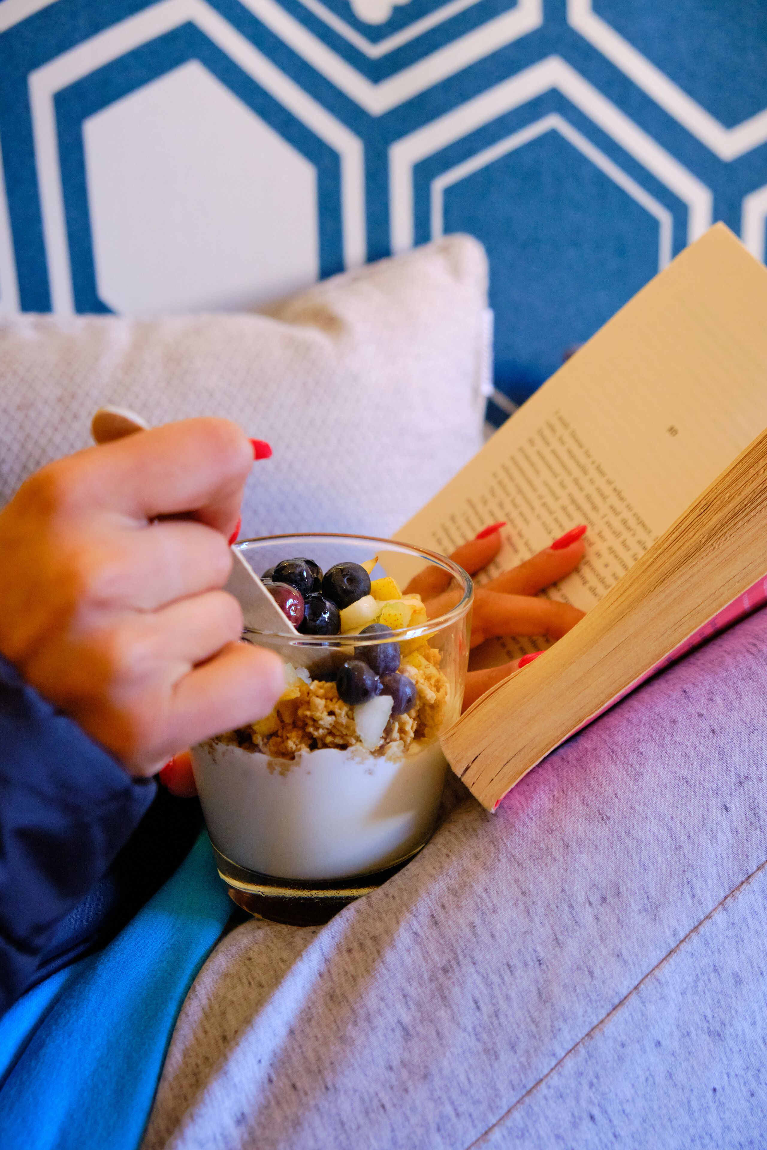 Uma pessoa comendo iogurte com frutas e granola, segurando uma colher, e lendo um livro.