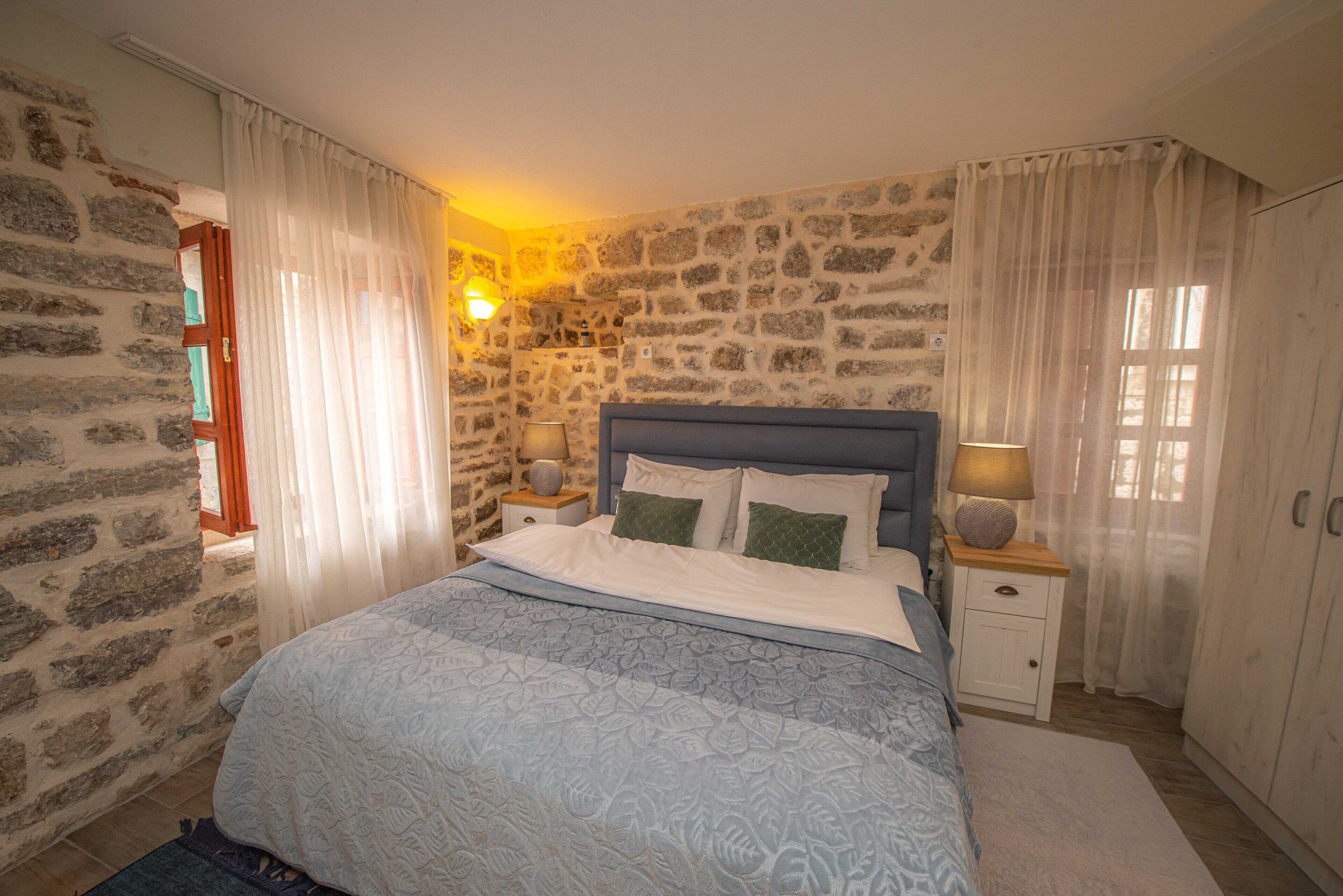 Quarto com parede de pedra, cama com colcha azul, almofadas brancas, mesa de cabeceira com abajur e um guarda-roupa.