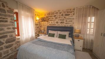 Quarto com parede de pedra, cama com colcha azul, almofadas brancas, mesa de cabeceira com abajur e um guarda-roupa.