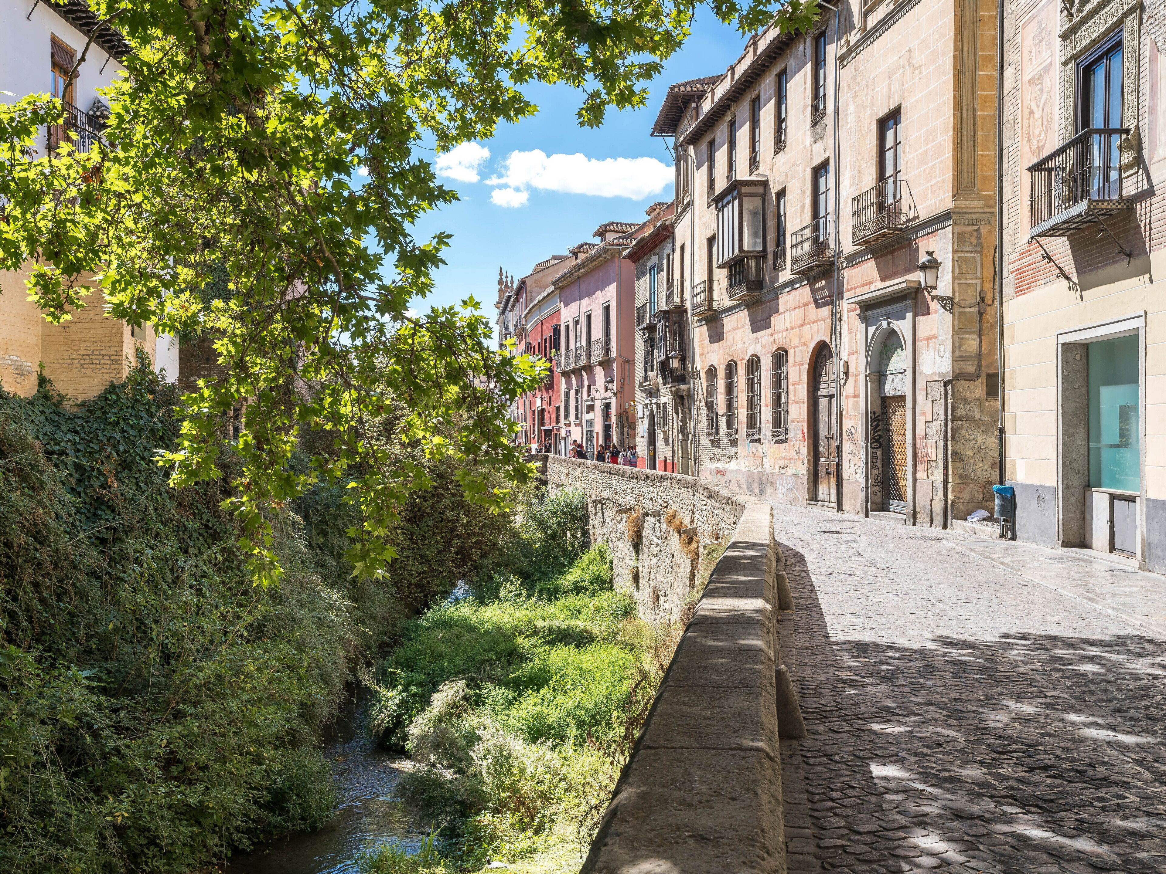 Rua de paralelepípedos ladeada por edifícios históricos, um pequeno canal e vegetação exuberante.