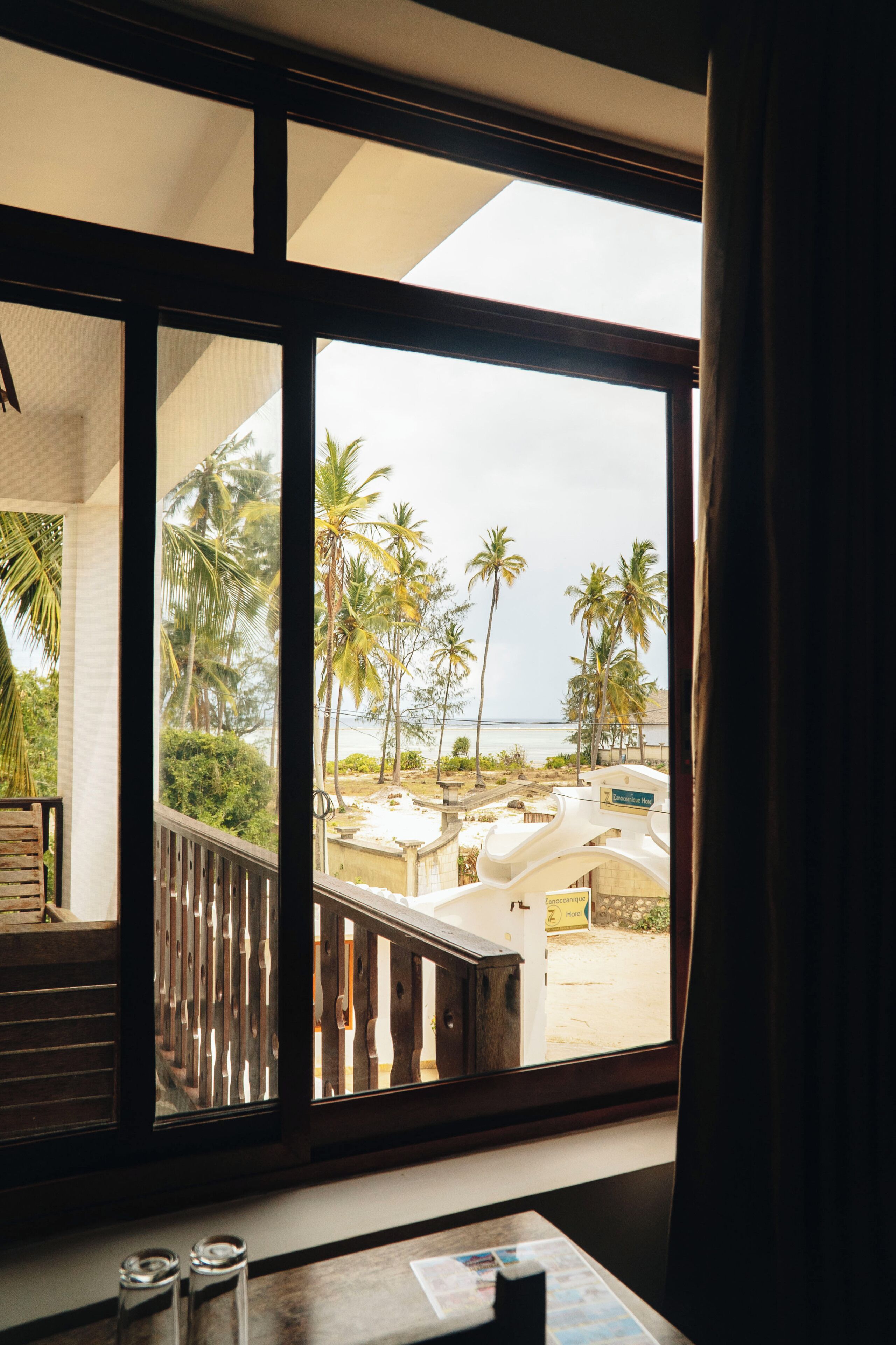Quarto com uma grande janela com vista para uma praia com palmeiras e uma piscina.