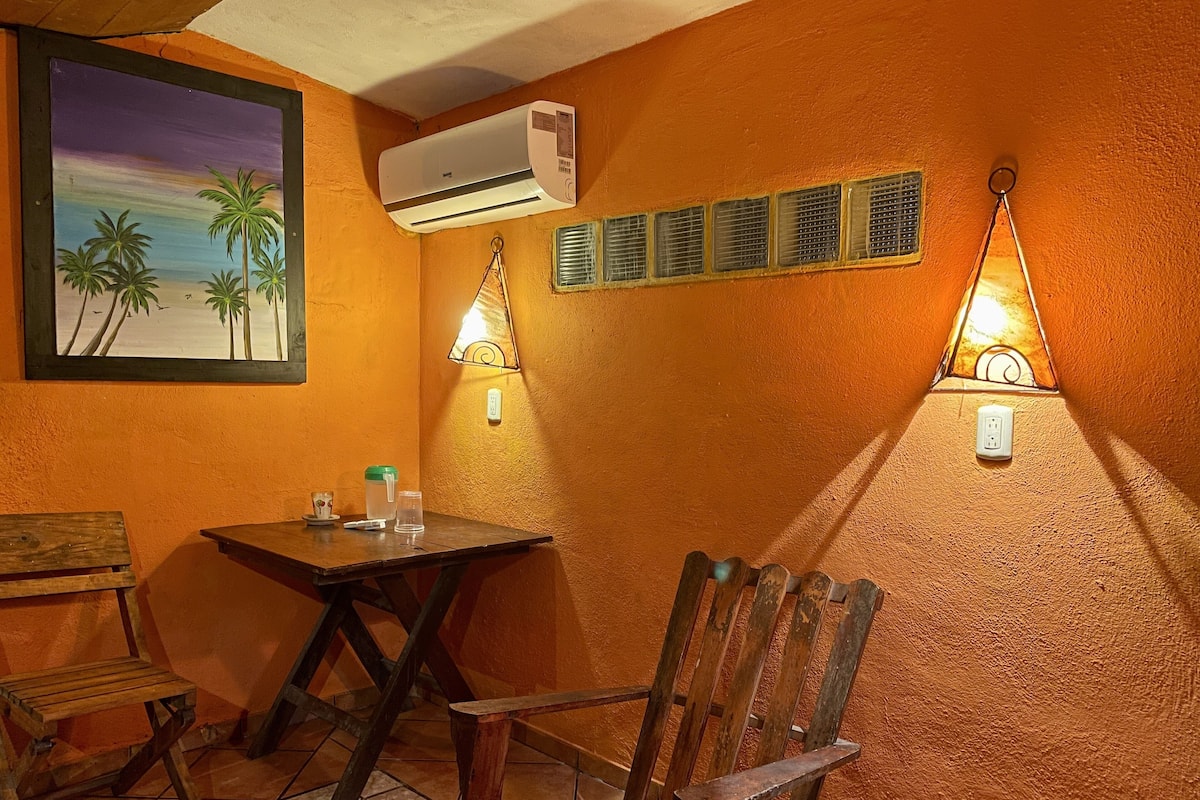 Quarto com uma mesa de madeira, uma cadeira, ar condicionado, pintura de palmeiras e duas luminárias na parede.