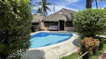 Piscina com estrutura de palha, rodeada por vegetação e palmeiras.