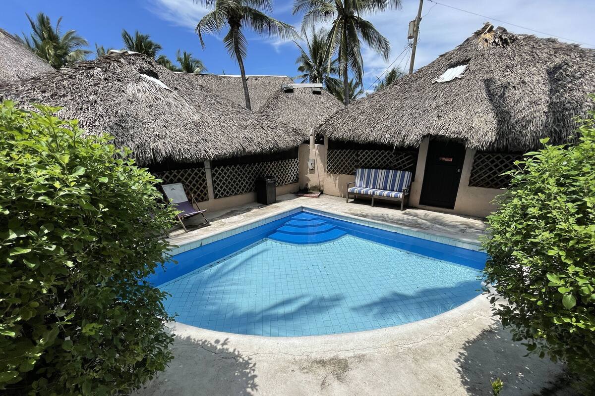 Uma piscina com deck revestido, cercada por construções de telhado de palha e vegetação exuberante.