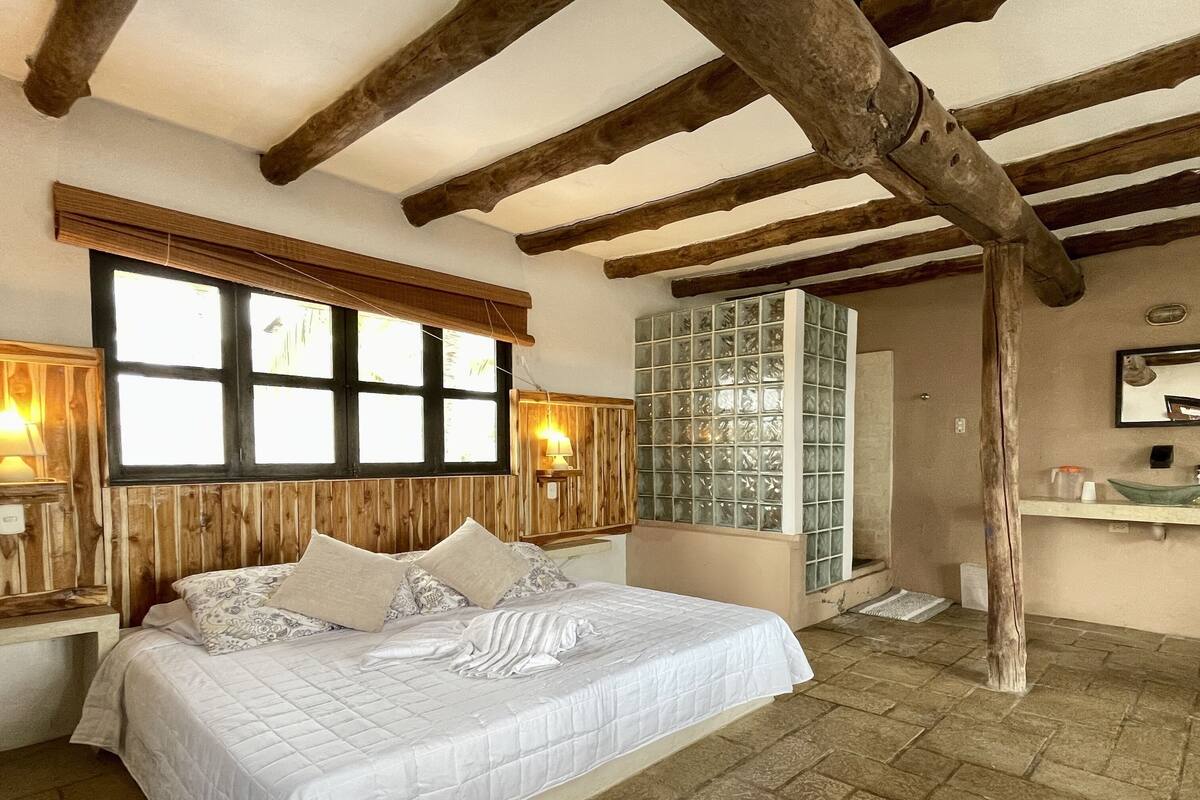 Quarto com uma cama grande, vigas de madeira, janela com persianas, luminária de cabeceira e banheiro com box de vidro.