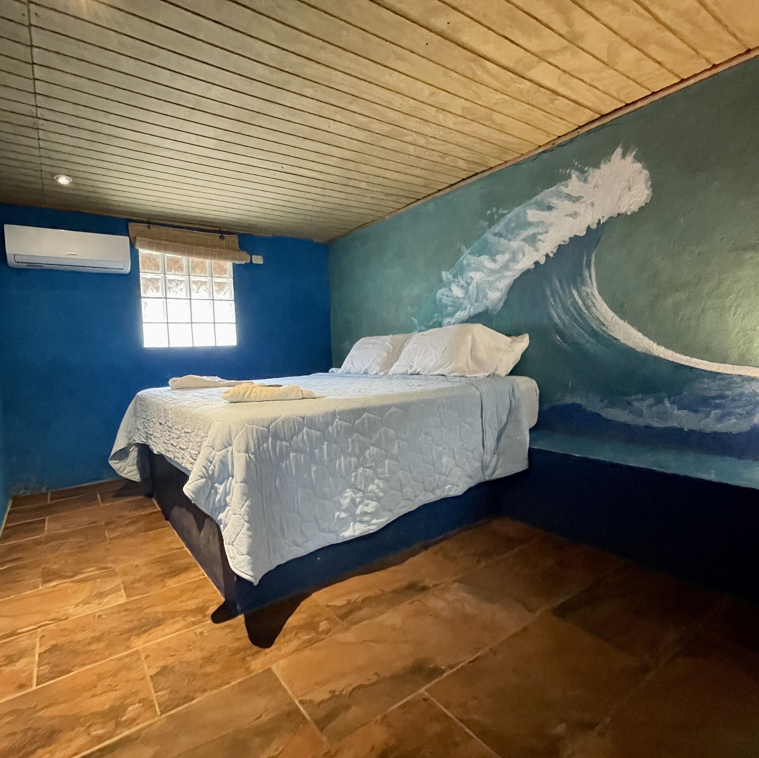 Quarto com uma cama, um mural de uma grande onda, uma janela e piso de azulejos.
