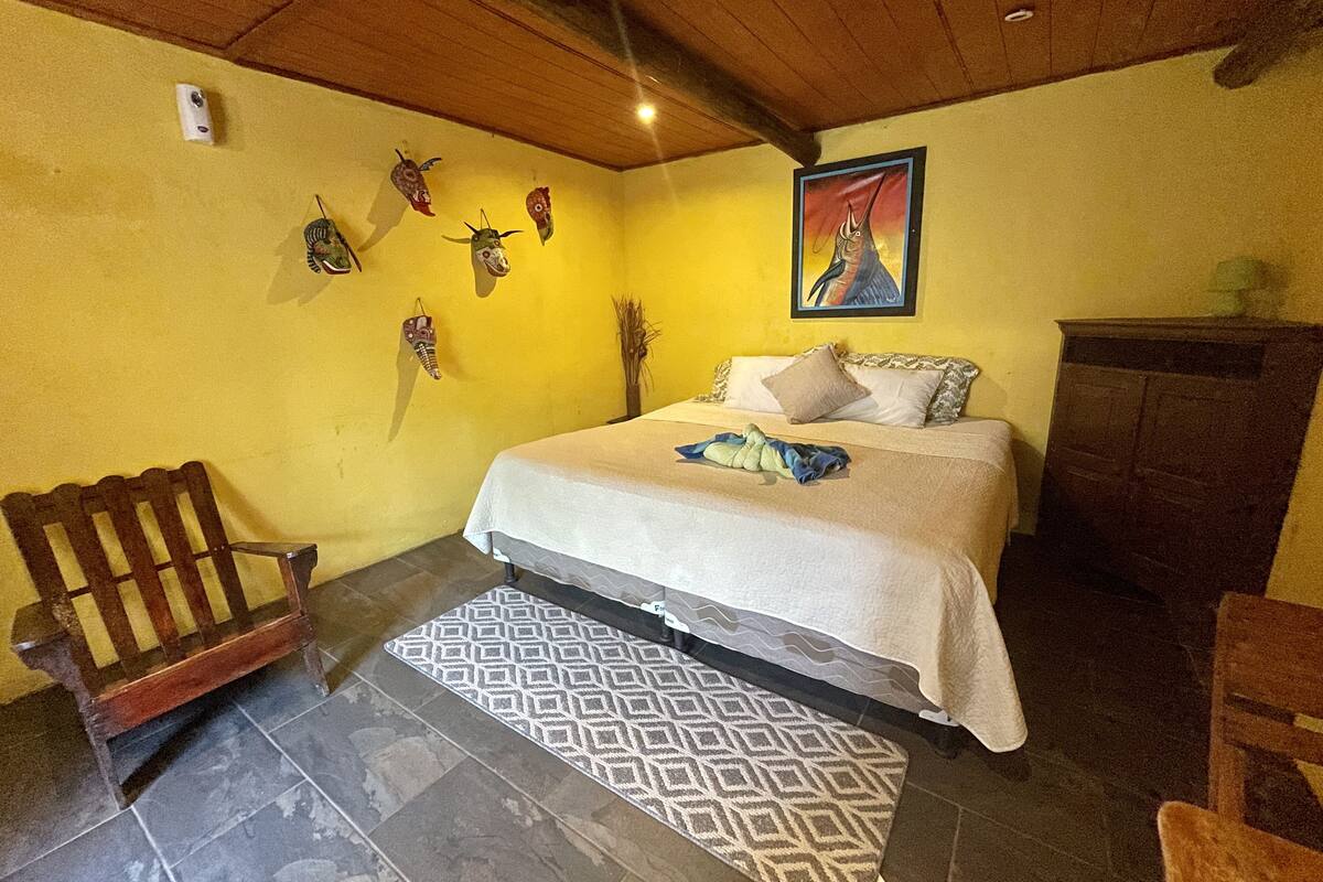 Quarto com cama, um banco de madeira, um tapete estampado e máscaras decorativas na parede.