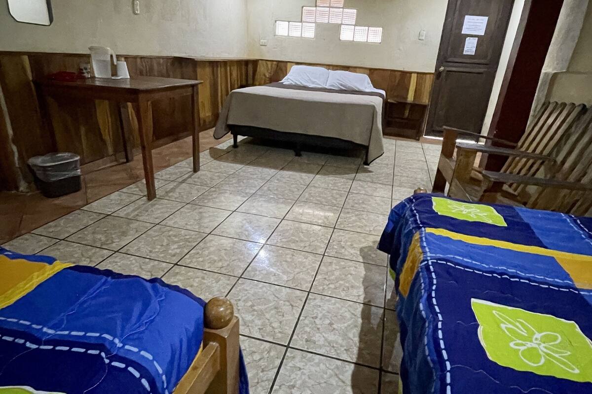 Quarto de hotel com duas camas de solteiro, uma escrivaninha de madeira, uma cadeira, um cesto de lixo pequeno e uma geladeira.