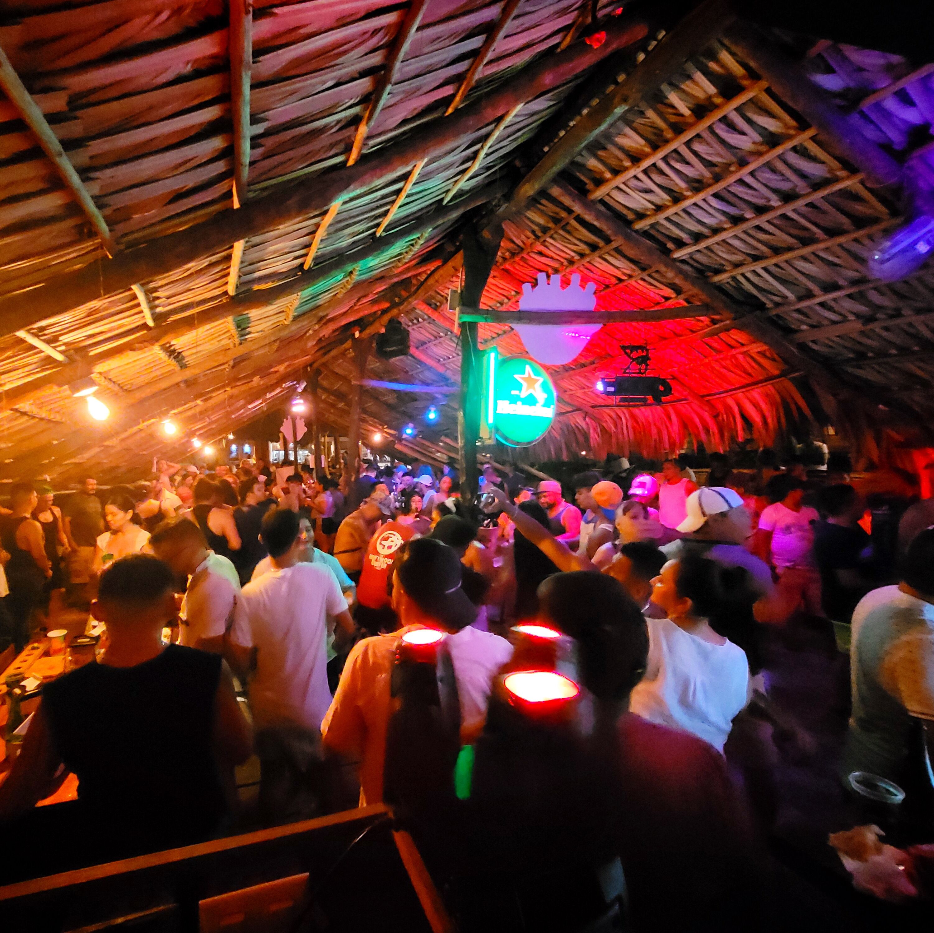 Um espaço interno cheio de gente, com telhado de palha e um letreiro de neon.