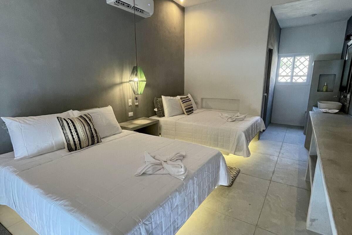 Quarto de hotel com duas camas, um ventilador de teto, uma pequena janela e um banheiro visível através de uma porta aberta.