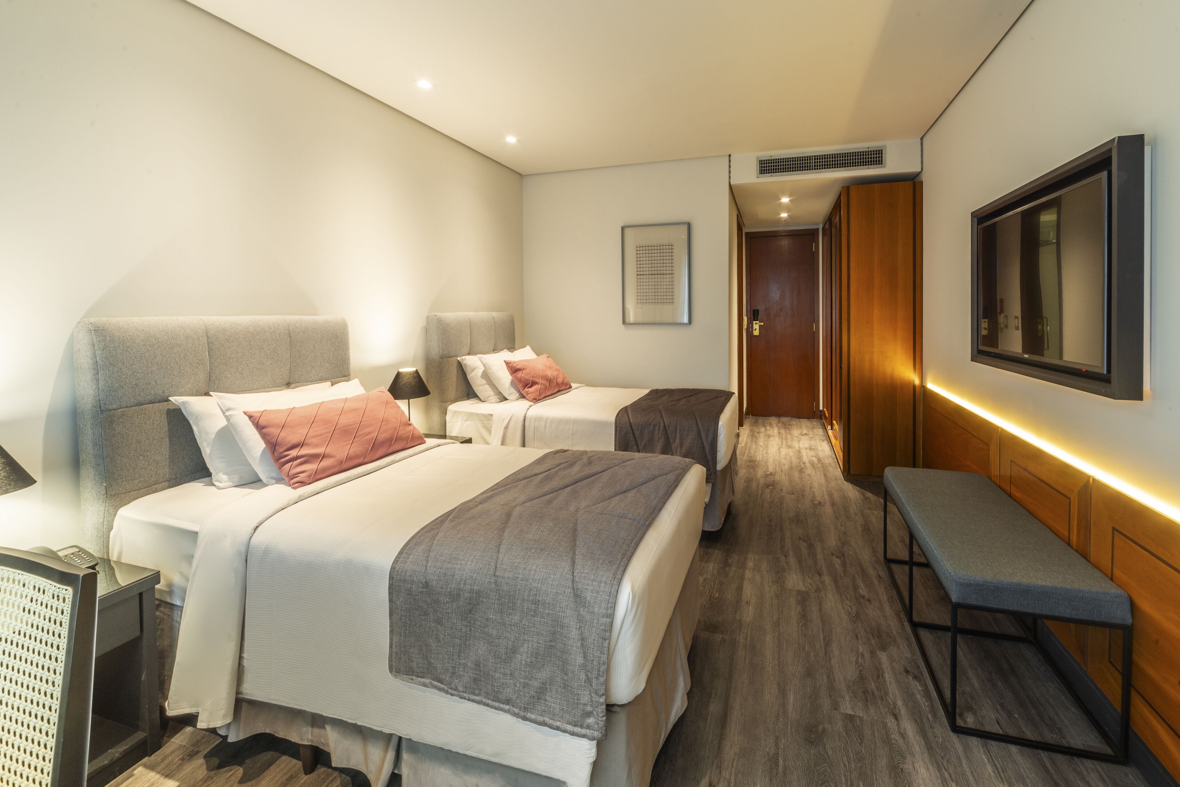 Quarto de hotel com duas camas, um banco, uma TV e um painel de parede de madeira.
