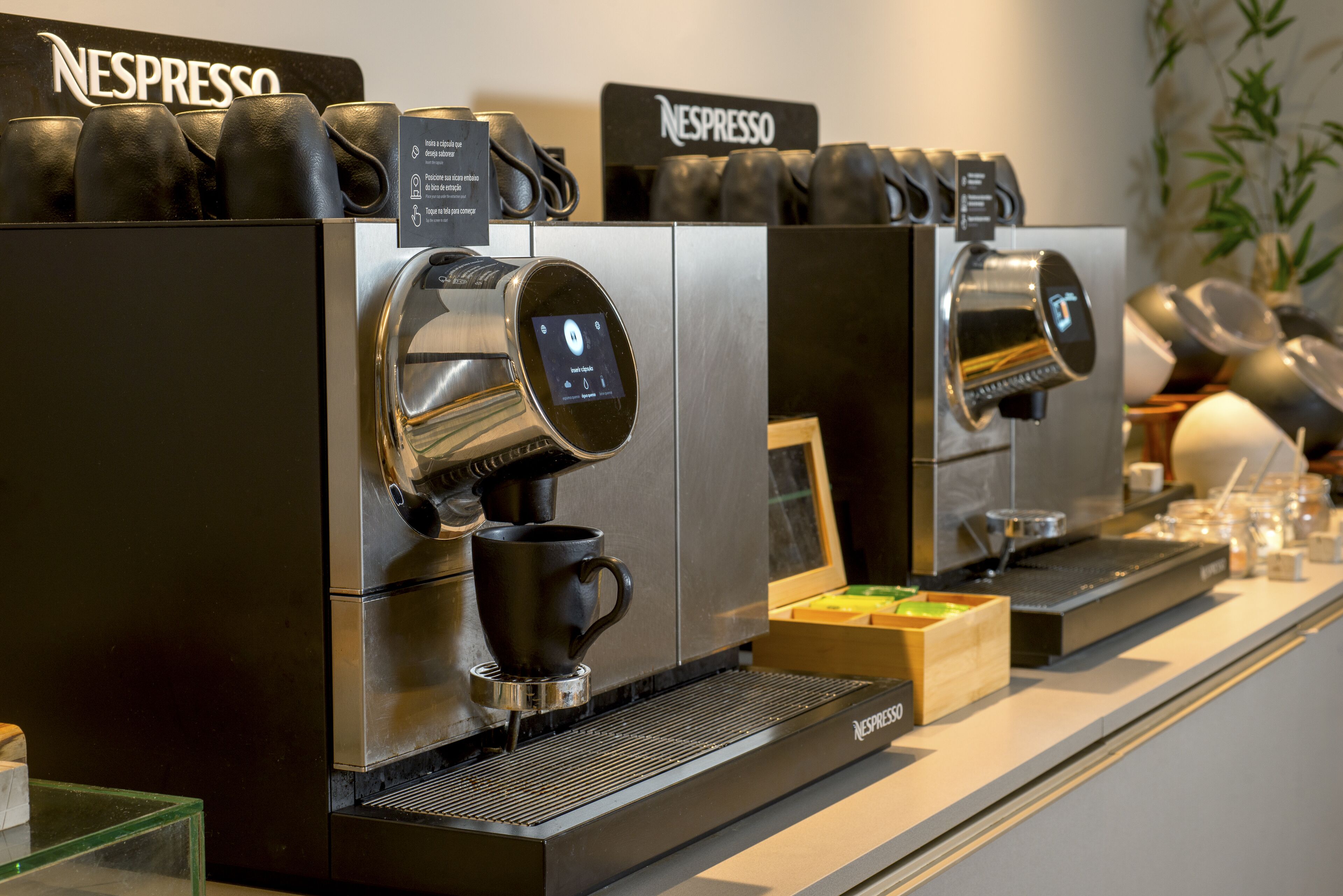 Máquina de café com visor digital, uma chávena de café e uma caixa de cápsulas de café.